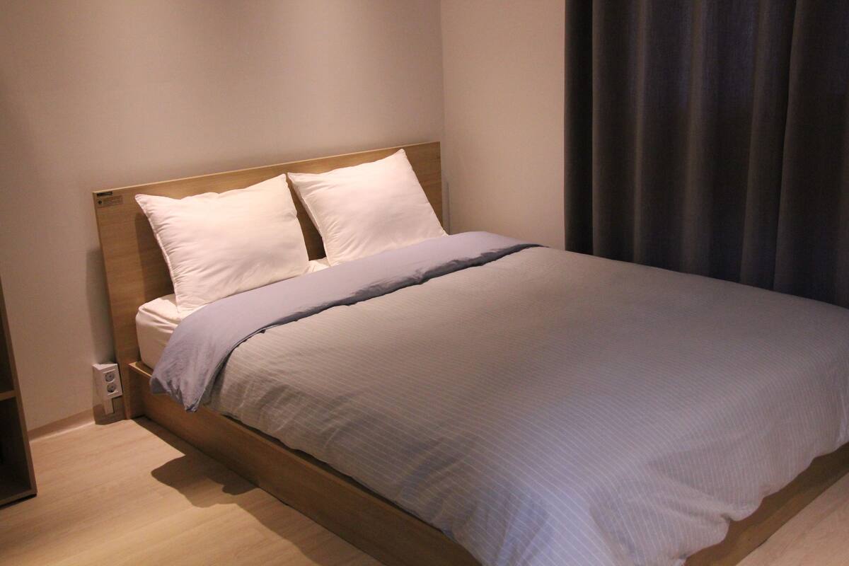 Cama bem arrumada com cabeceira de madeira, uma mesa de cabeceira e parede lisa com cortina.