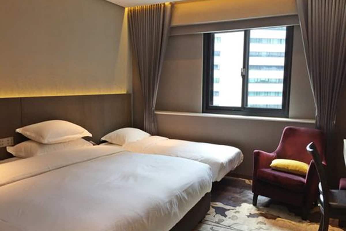 Quarto de hotel com duas camas, uma poltrona vermelha, uma janela grande com cortinas e vista para a cidade.