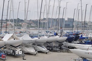 Uma marina com vários veleiros atracados, uma pessoa perto dos barcos e edifícios ao fundo.