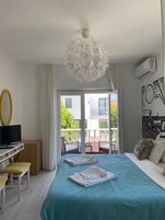 Quarto de hotel com cama, escrivaninha com computador, televisão, varanda com cadeiras e vista para o exterior.