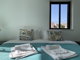 Quarto de hotel com uma cama, dois travesseiros estampados, uma janela com vista e duas toalhas dobradas sobre a cama.