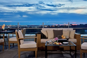 Um terraço com móveis de vime, uma mesa de vidro e vista para o horizonte urbano ao entardecer.