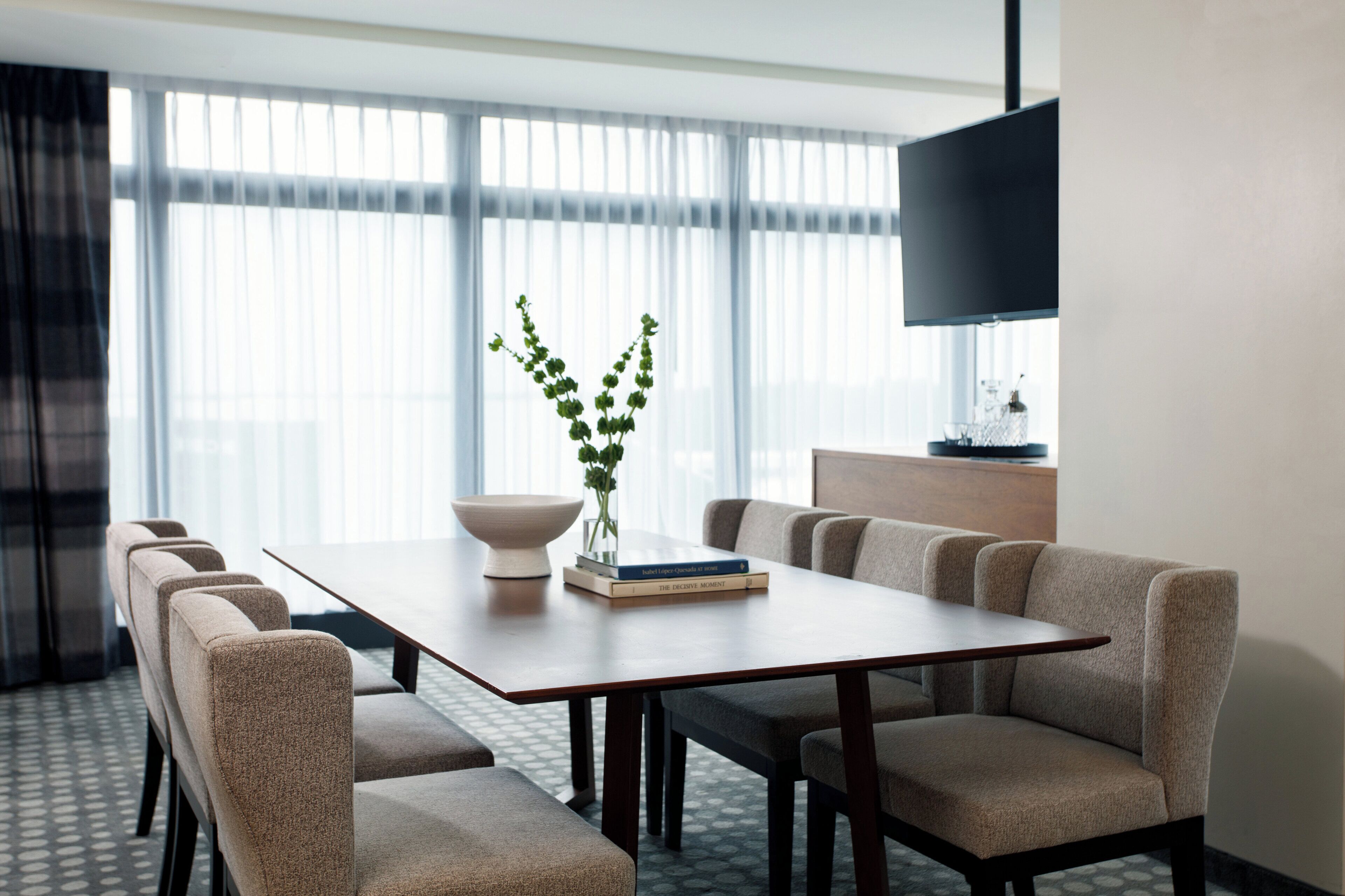 Uma área de refeições de hotel com uma mesa retangular, seis cadeiras estofadas, uma tigela branca, dois livros e um vaso com folhagem verde.