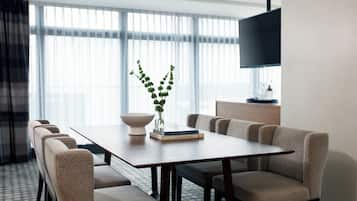Uma área de refeições de hotel com uma mesa retangular, seis cadeiras estofadas, uma tigela branca, dois livros e um vaso com folhagem verde.