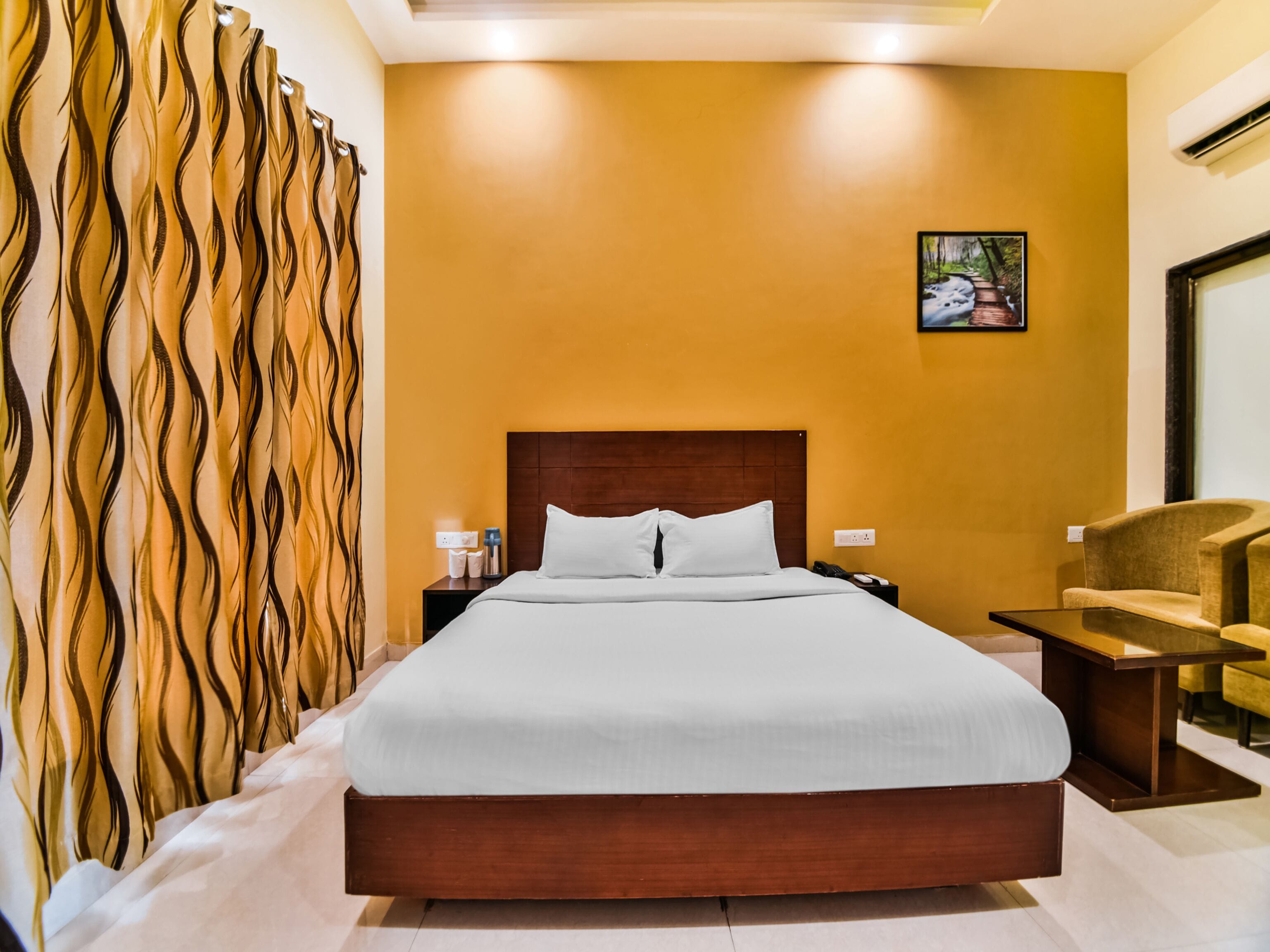 Quarto de hotel com uma cama grande, cabeceira de madeira, mesa de cabeceira, uma cadeira e um quadro na parede.