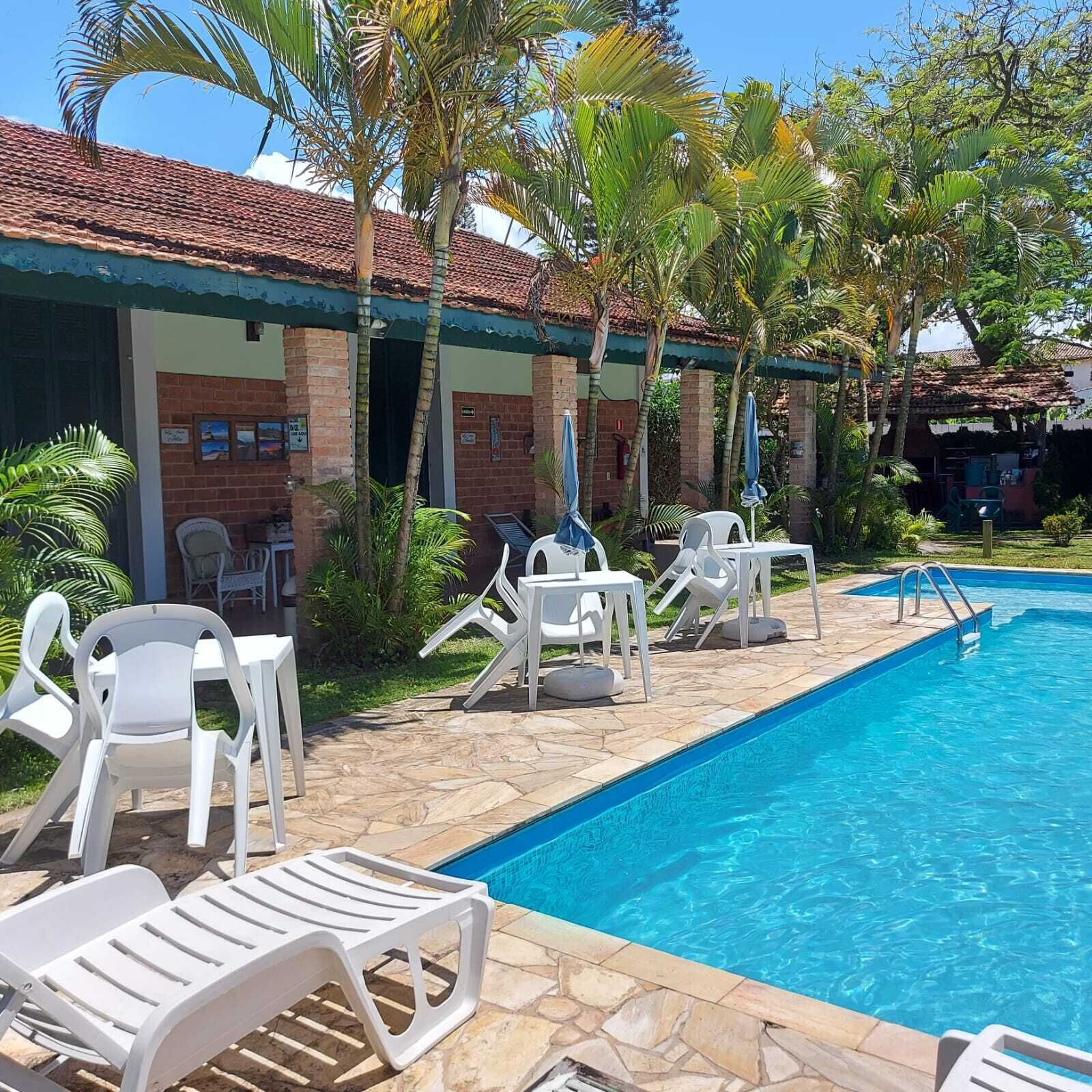 Piscina com cadeiras de sol brancas e guarda-sóis, cercada por palmeiras e um edifício com telhado em azulejos.