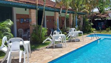 Piscina com cadeiras de sol brancas e guarda-sóis, cercada por palmeiras e um edifício com telhado em azulejos.