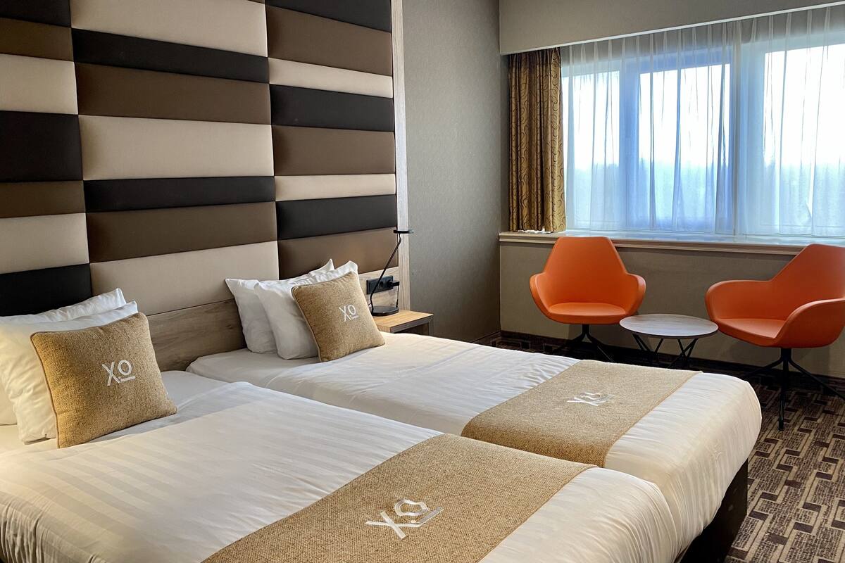 Quarto de hotel com uma cama grande, duas almofadas, uma mesinha de cabeceira, uma cadeira e uma janela com cortinas.