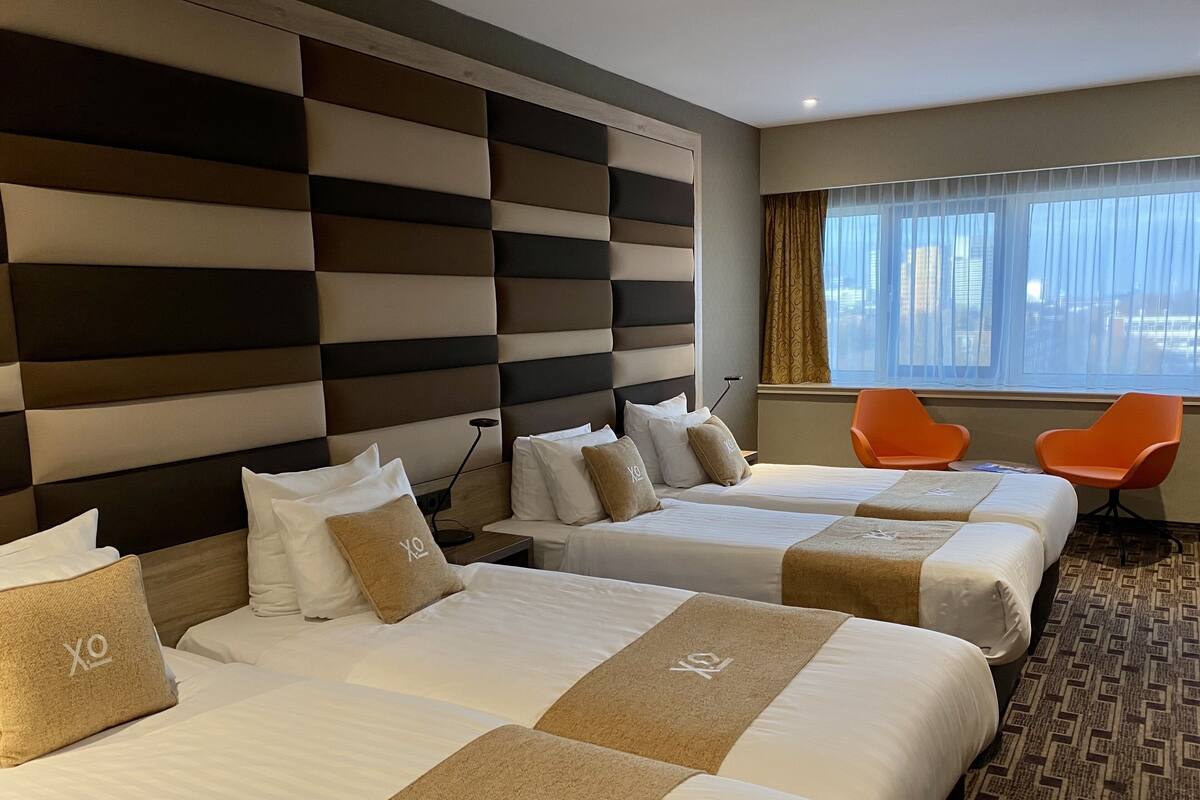Quarto de hotel com duas camas, uma janela grande com cortinas e duas cadeiras laranja.