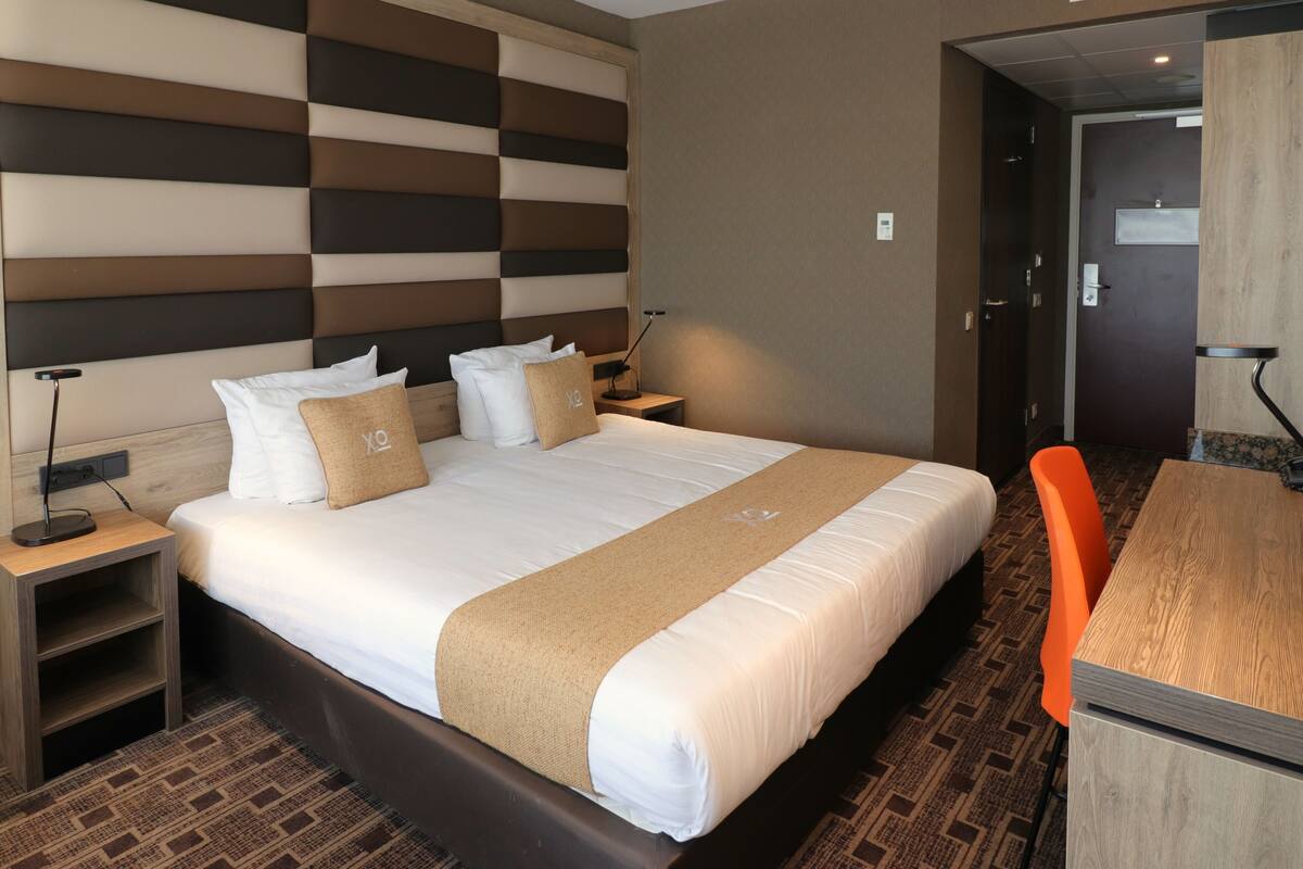 Quarto de hotel com uma cama grande, cabeceira de madeira, mesa de cabeceira com abajur, prateleira pequena e escrivaninha de madeira com uma cadeira.