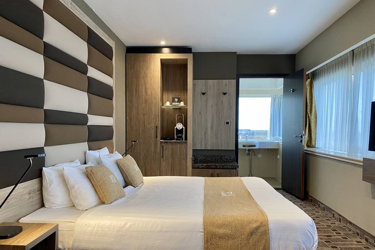 Quarto de hotel moderno com uma cama grande, um guarda-roupa de madeira, um banheiro pequeno e vista para a cidade.