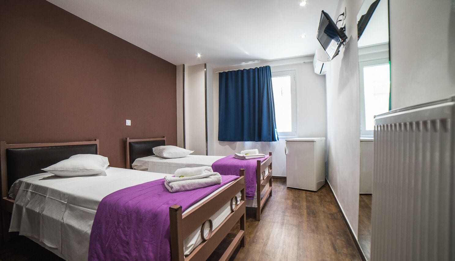 Quarto de hotel com duas camas, um pequeno aquecedor, uma janela com cortinas azuis e uma televisão fixada na parede.