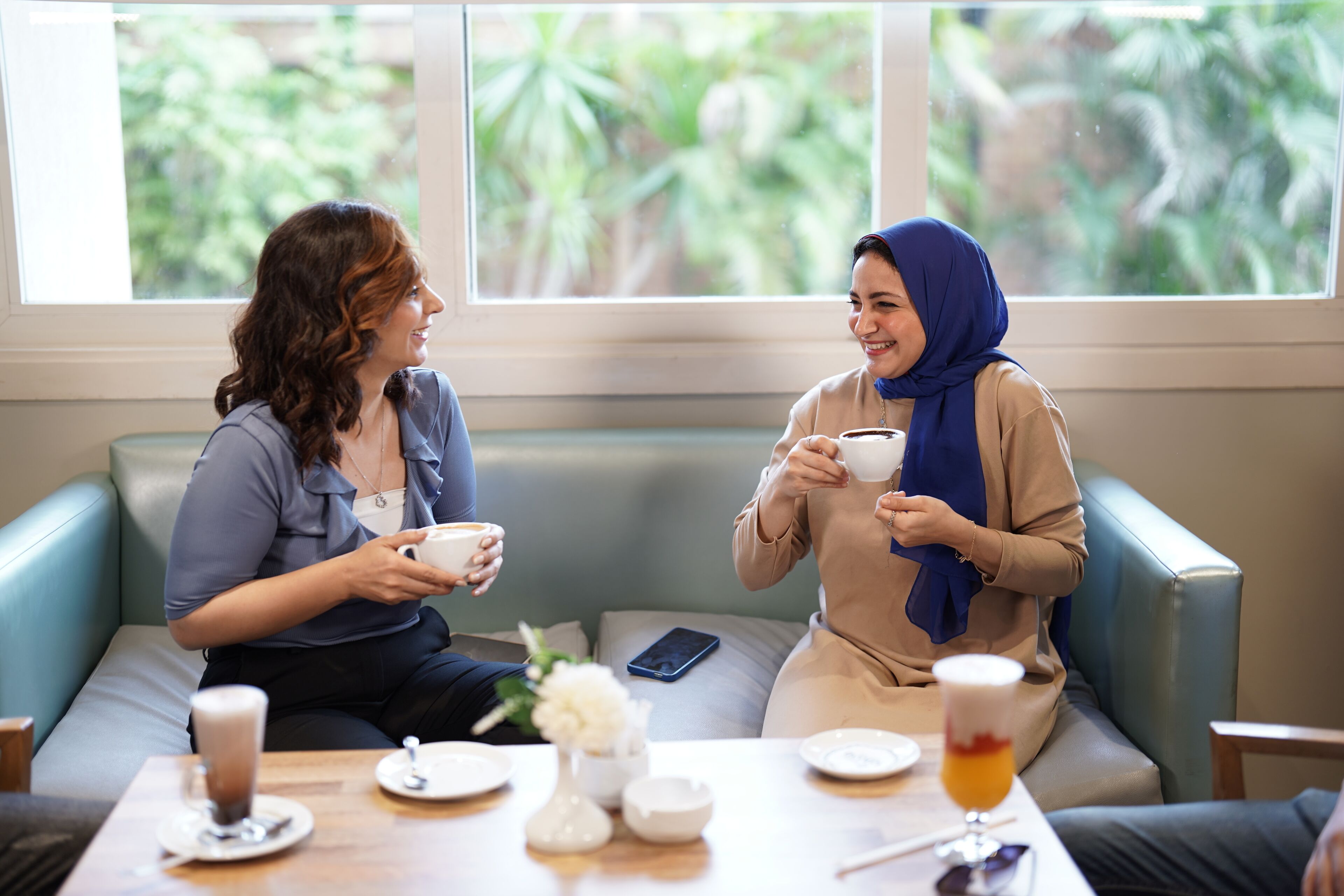 Duas pessoas sentadas em um sofá, segurando xícaras, com uma mesa à frente que possui pratos, um vaso com flores e um copo de suco.