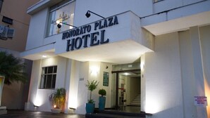 Exterior do Hotel Honorato Plaza, com uma placa acima da entrada, uma escadaria que leva para o interior e duas plantas em vasos à frente.
