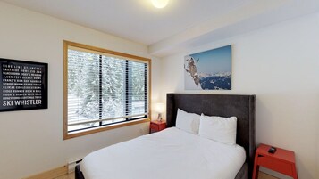Um quarto com uma cama grande, uma mesinha de cabeceira, uma janela com persianas, um quadro na parede e um letreiro listando pistas de esqui.