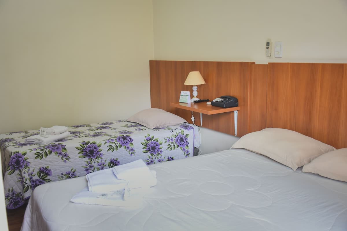 Quarto de hotel com duas camas, cada uma com colcha florida, cabeceira de madeira, mesa de cabeceira com luminária, telefone e um aviso indicando os serviços do quarto.