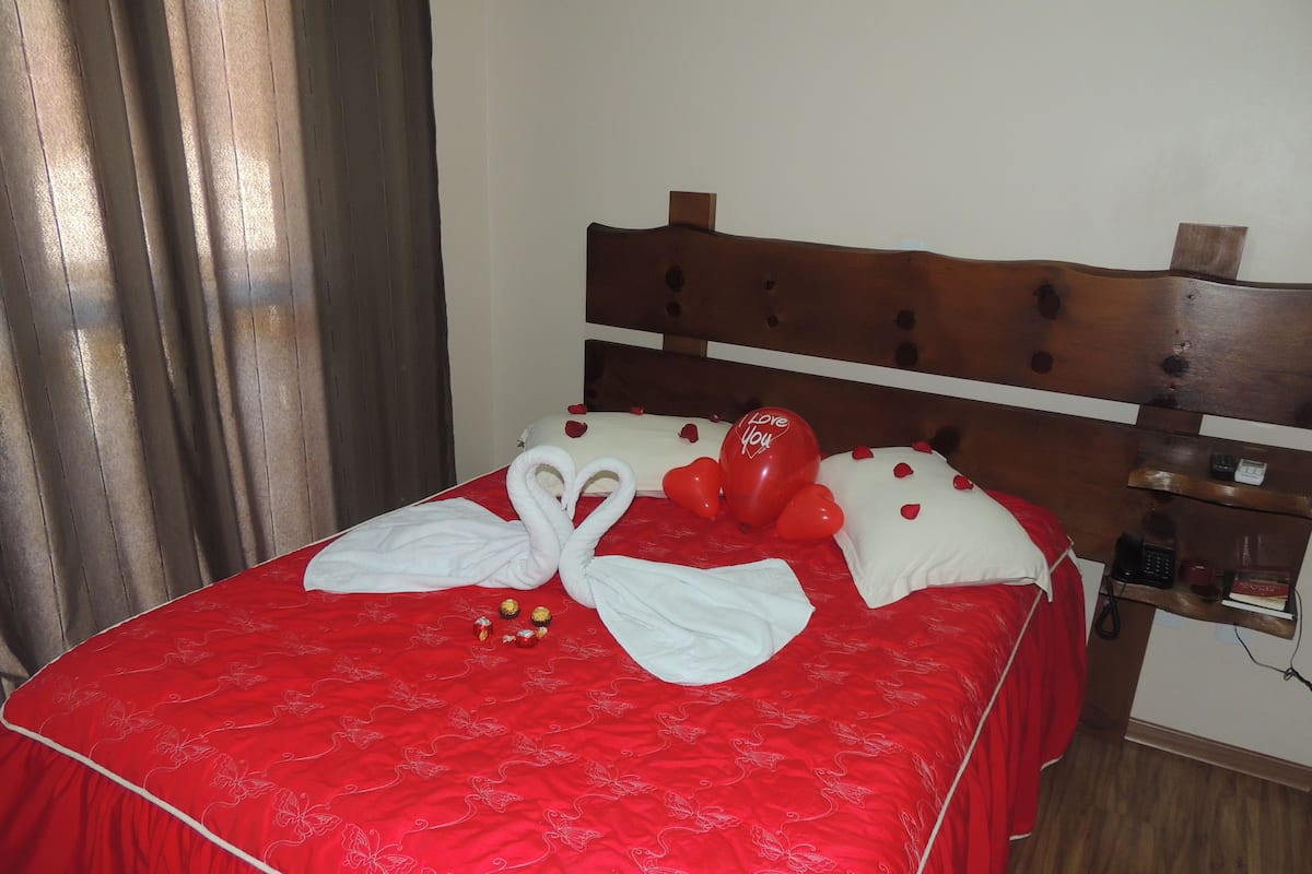Uma cama com colcha vermelha, duas toalhas brancas dispostas em formato de cisnes, um balão em formato de coração vermelho e uma almofada branca com rosas vermelhas.