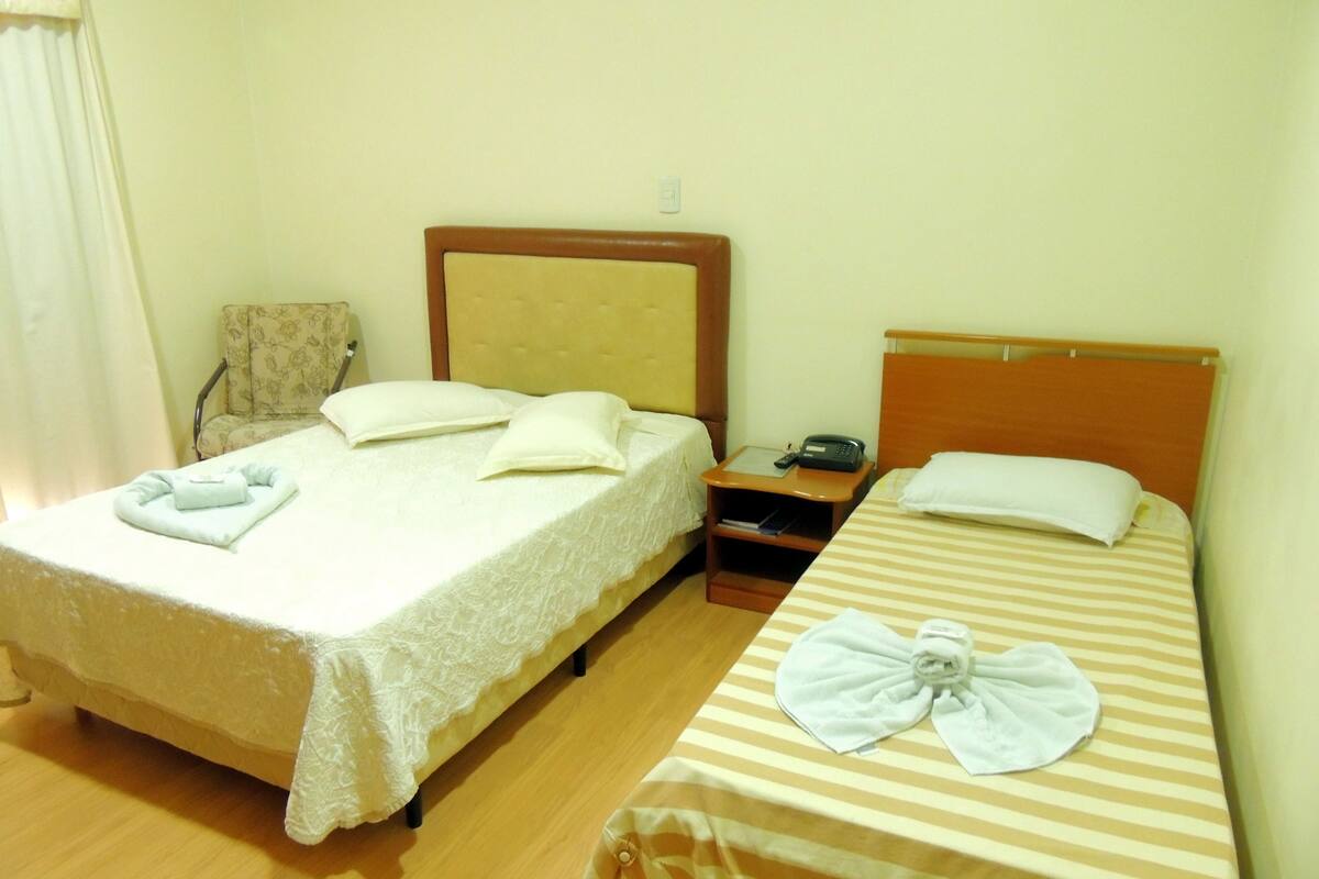 Quarto de hotel com duas camas, uma mesa de cabeceira com telefone e uma cadeira.