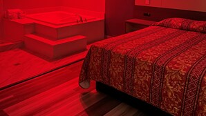 Quarto com banheira iluminada em vermelho, cama com colcha estampada e piso de madeira.