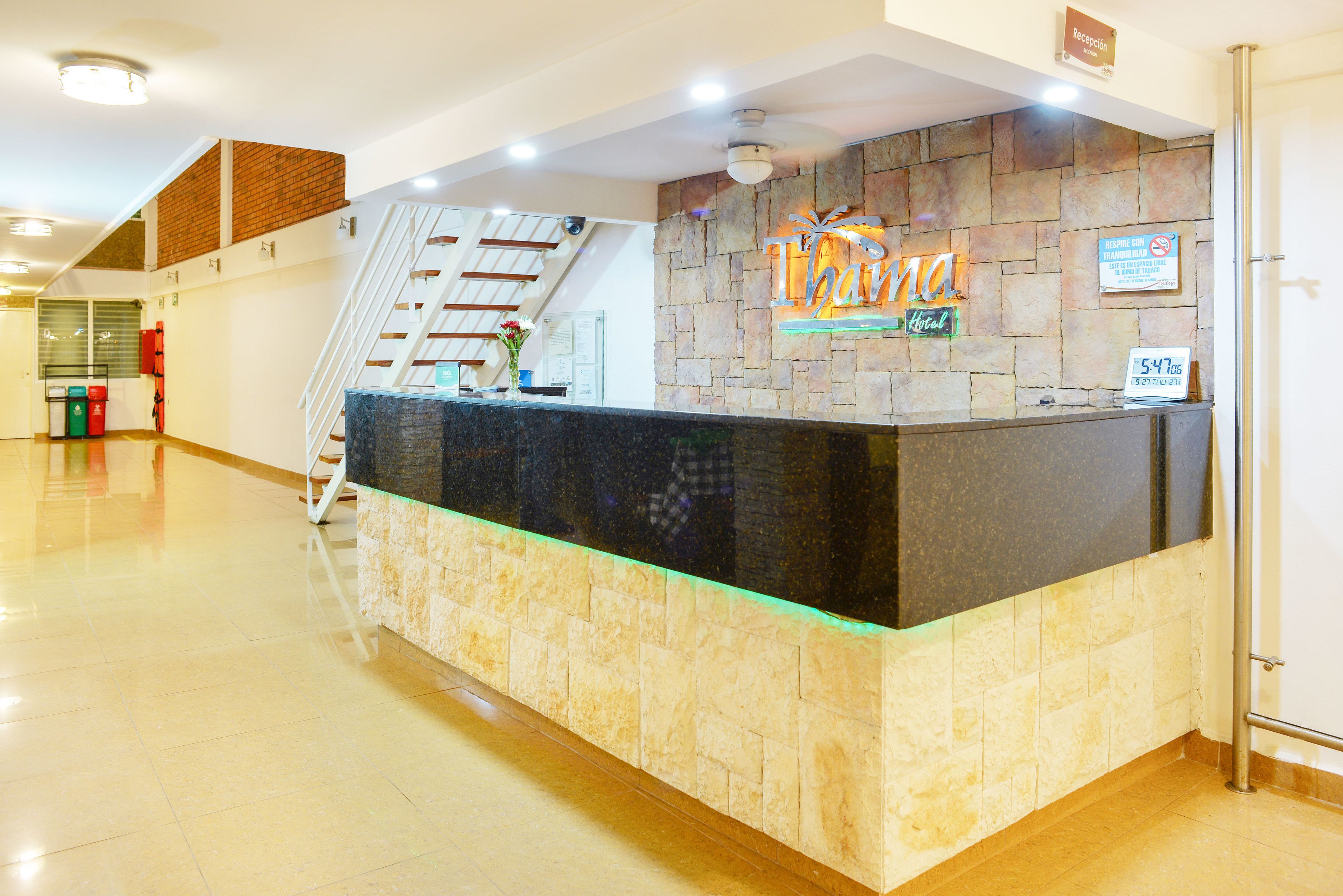 Uma receção com parede de pedra ao fundo, um letreiro com a inscrição "Hana Hotel" e uma escadaria à esquerda.