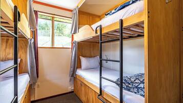 Quarto pequeno e bem iluminado, com beliches, janela com cortinas e porta com ganchos.