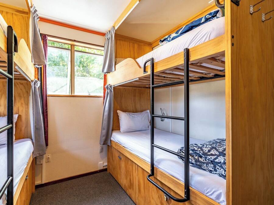 Quarto pequeno e bem iluminado, com beliches, janela com cortinas e porta com ganchos.