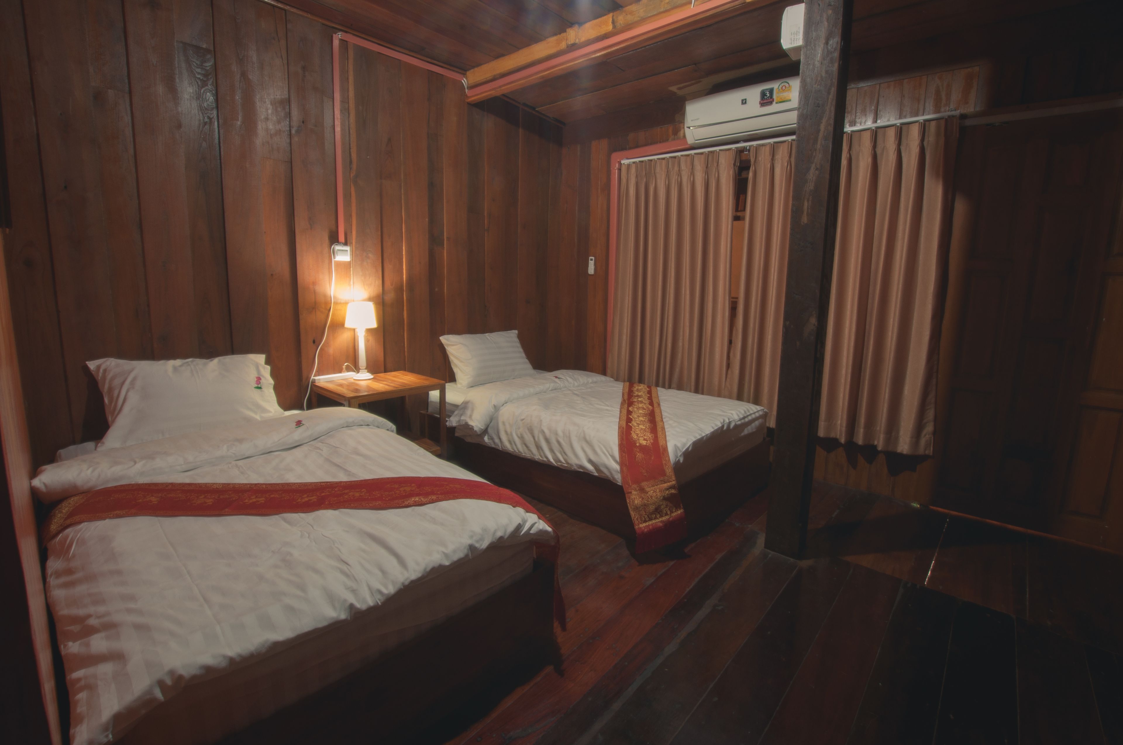 Quarto com duas camas, uma mesa de cabeceira com abajur, paredes de madeira e cortina.