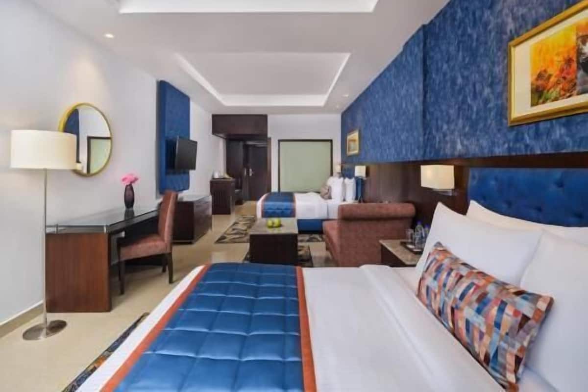 Quarto de hotel com uma cama, escrivaninha, poltrona, sofá, televisão e um quadro na parede.