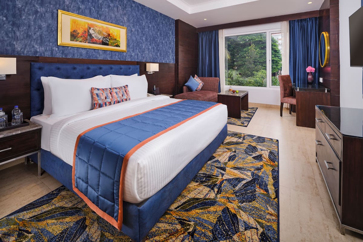 Quarto de hotel com uma cama grande, uma escrivaninha, uma cadeira, uma televisão e uma janela com vista para vegetação.