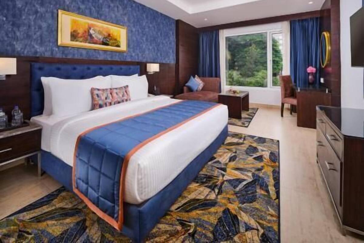 Quarto de hotel com uma cama grande, cabeceira, mesa de cabeceira, escrivaninha, cadeira, cômoda e janela com cortinas.