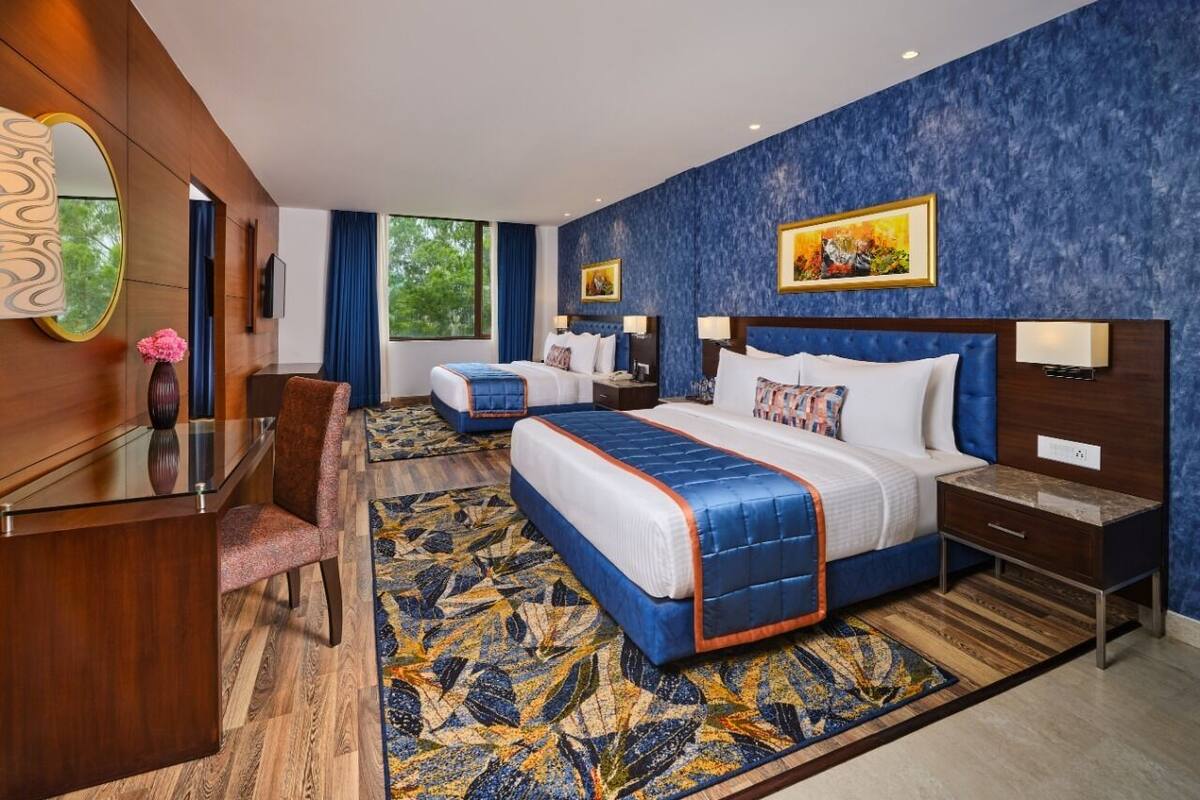 Quarto de hotel com uma cama grande, uma cama menor, uma mesa com cadeira e uma janela ampla.