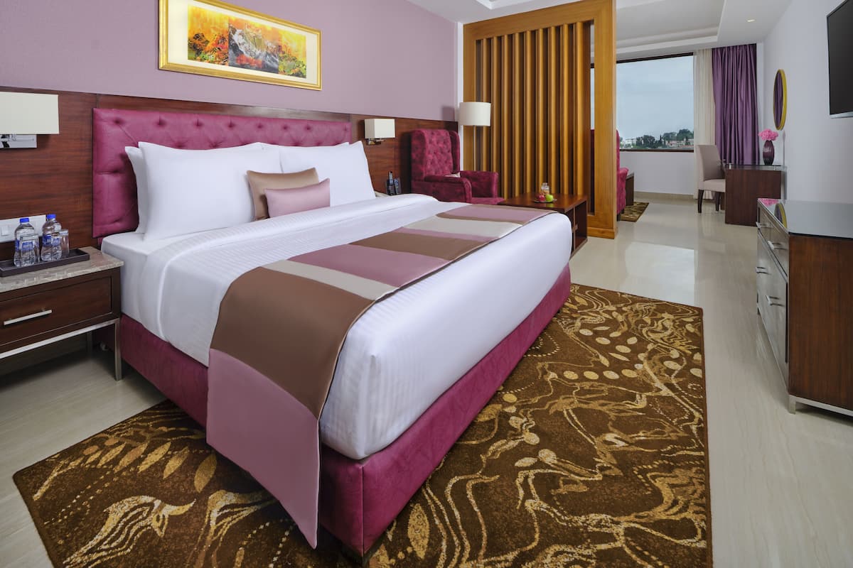 Quarto de hotel com cama grande, mesa de cabeceira com garrafas de água, cabeceira de madeira, grande janela com cortinas, escrivaninha e uma cadeira.