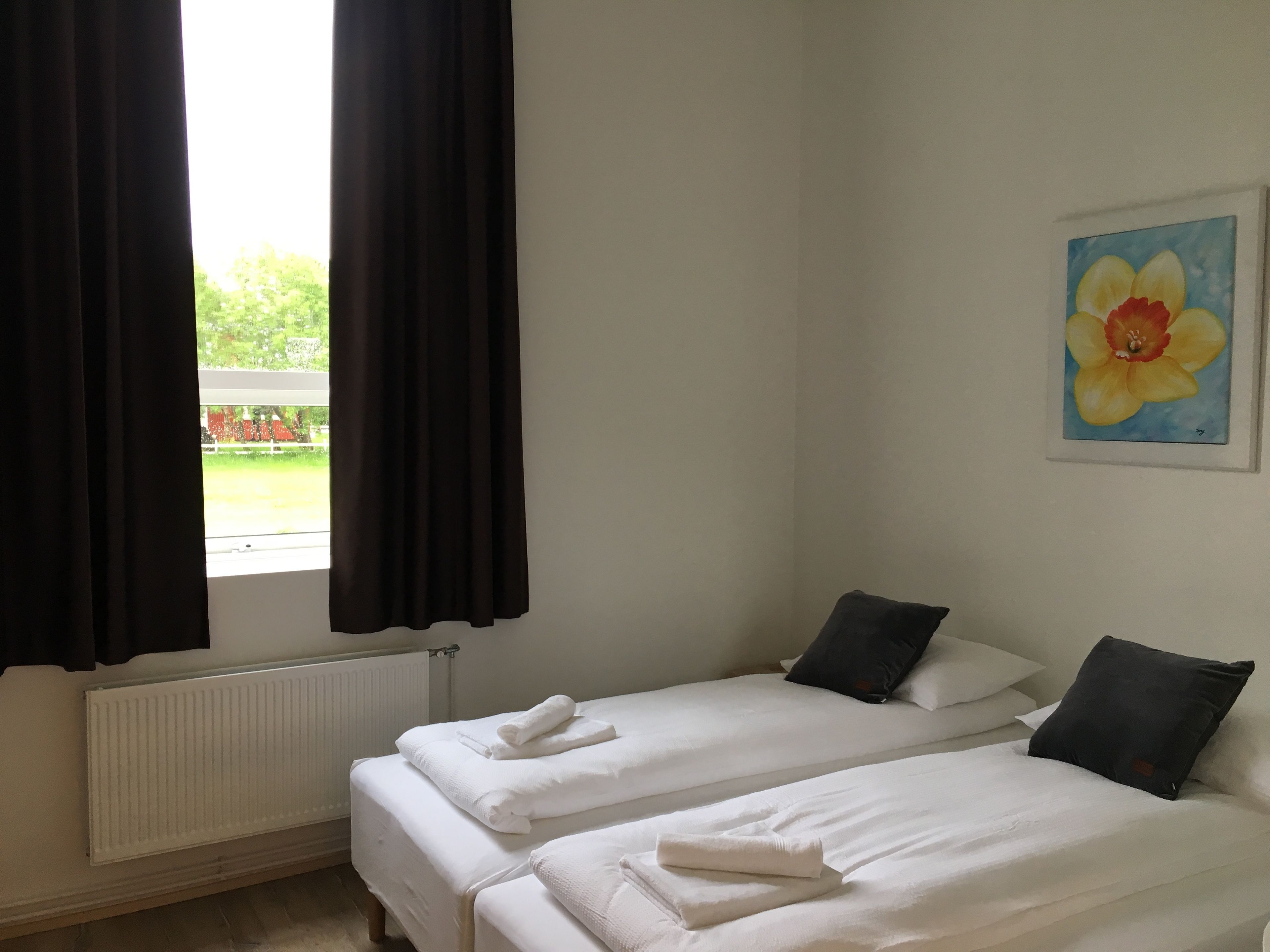 Quarto com duas camas, roupa de cama branca, cortinas pretas, radiador e um quadro de uma flor na parede.