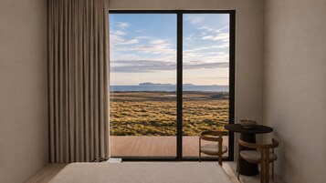 Quarto com uma grande porta de correr de vidro que dá para uma paisagem gramada e montanhas ao longe. Há uma pequena mesa redonda com uma cesta de frutas e duas cadeiras.