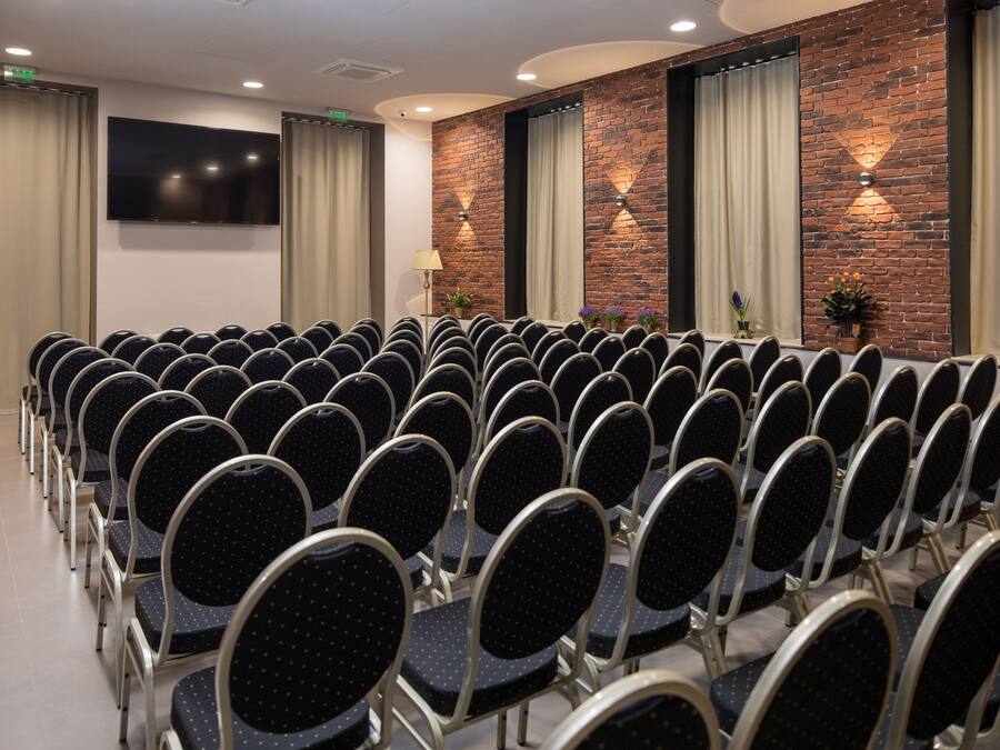Sala de conferências com fileiras de cadeiras, uma TV de tela plana, parede de tijolos e cortinas.