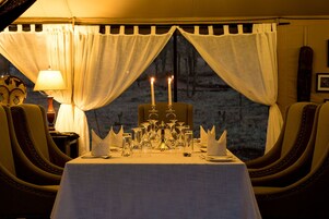 Mesa de jantar posta para dois, com velas, um abajur e vista para uma paisagem nevada através de cortinas translúcidas.