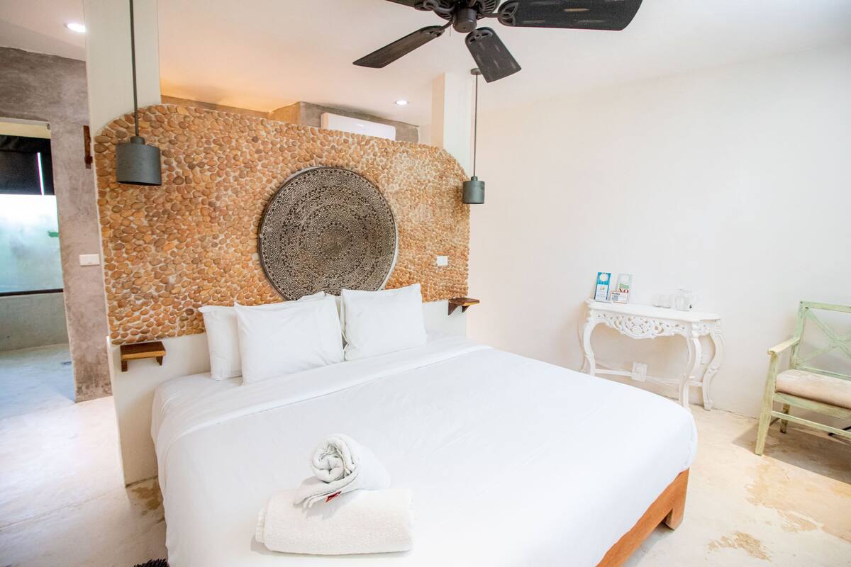 Quarto de hotel com uma cama grande, parede de pedra, objeto decorativo circular na parede, ventilador de teto, mesa pequena com produtos de higiene pessoal e uma cadeira.
