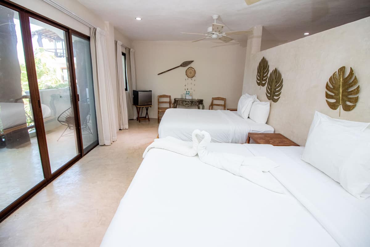 Quarto de hotel com duas camas, televisão, mesa pequena e varanda com cadeira.