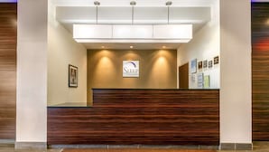 Uma área de recepção de hotel com um balcão de madeira, uma placa com o nome 'Sleep Inns & Suites' e um tapete de boas-vindas com a inscrição 'welcome. Columbia'.