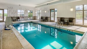 Piscina interna com água cristalina, cercada por cadeiras de lounge e mesas, grandes janelas e um teto com difusores de ar condicionado.