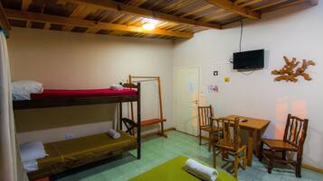 Um quarto com beliches, um colchonete verde, cadeiras de madeira, uma mesa, uma televisão e uma porta.
