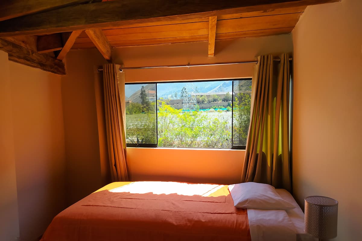 Quarto com uma janela grande, vigas de madeira no teto e uma cama com cobertor vermelho.