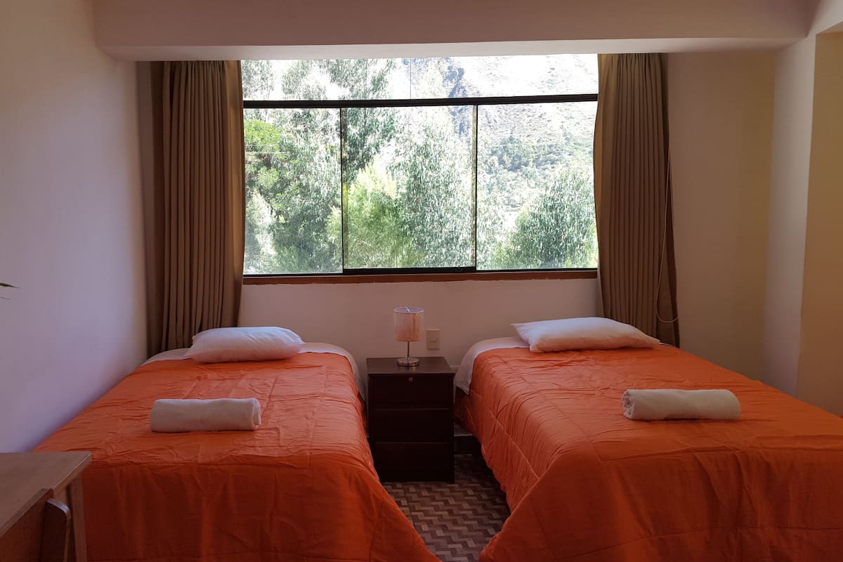 Quarto com duas camas, uma mesa de cabeceira, uma luminária, um vaso com planta e uma janela com cortinas.
