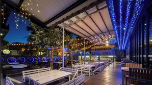 Uma área de refeições ao ar livre com mesas e bancos de madeira, luzes de fada e uma seção coberta com iluminação azul ambiente.