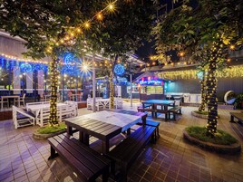 Uma área de refeições ao ar livre com mesas e bancos de madeira, luzes de fada e um restaurante chamado 'Bler Garden' ao fundo.