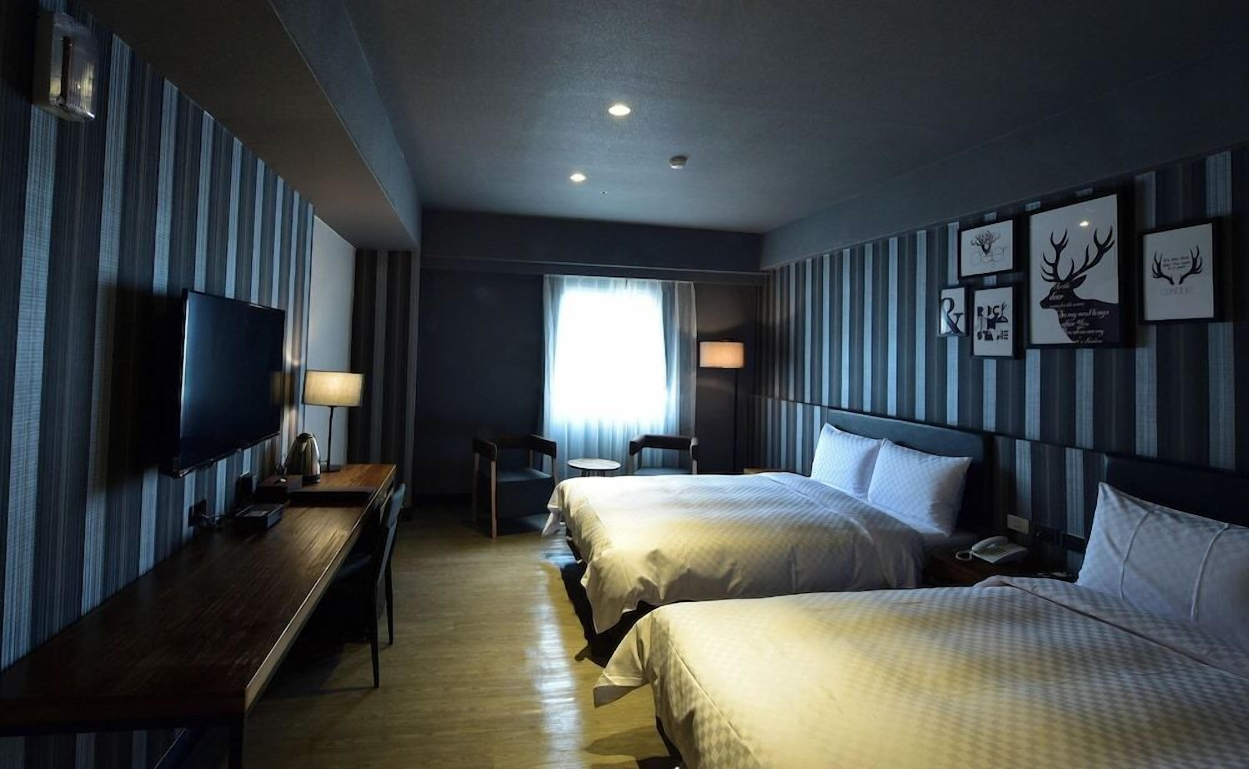 Quarto de hotel com duas camas, uma escrivaninha, televisão e janela com cortinas.