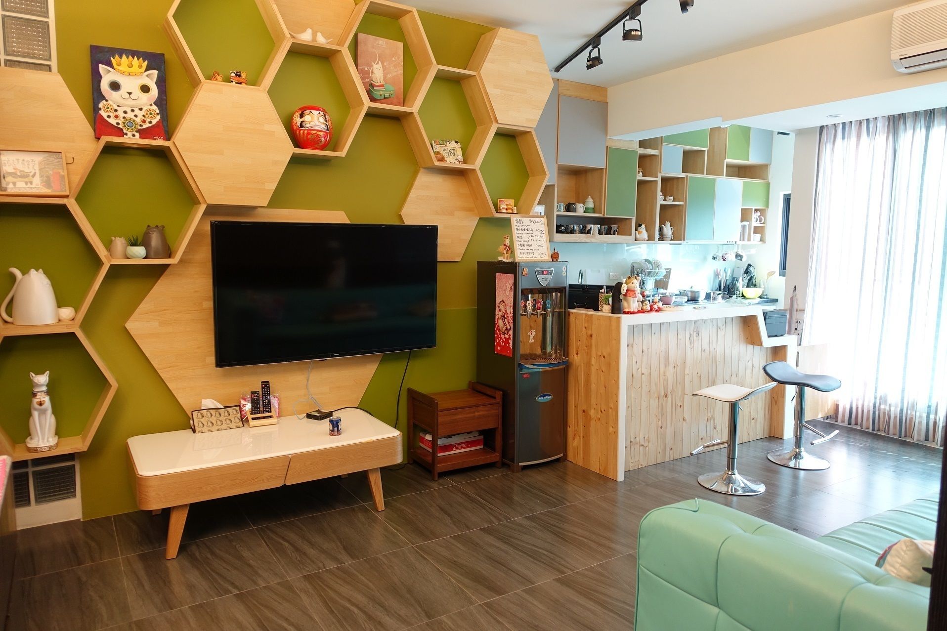 Uma sala de estar moderna com uma grande TV de tela plana, prateleiras hexagonais de madeira, um sofá verde e uma área de cozinha com banquetas.