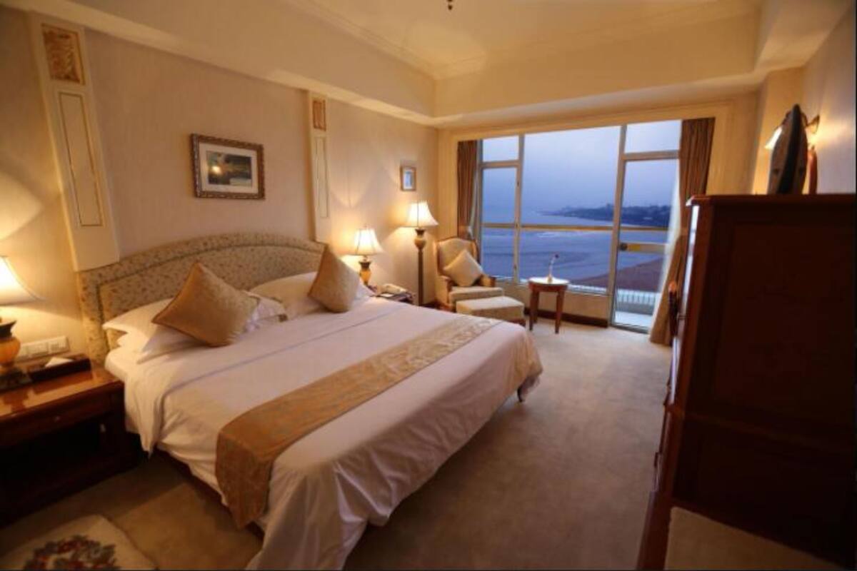 Quarto de hotel com uma cama grande, duas mesas de cabeceira, uma cadeira, uma mesa pequena e uma janela ampla com vista para o mar.