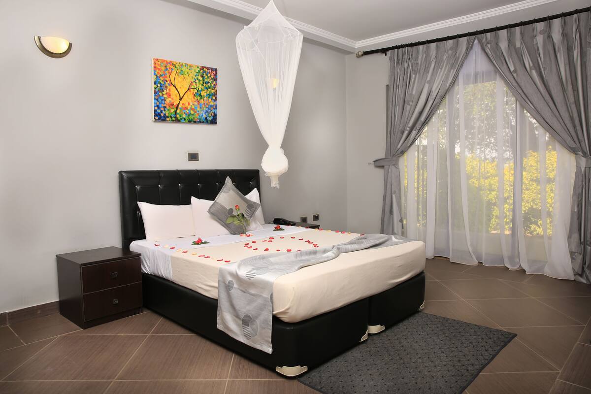 Uma cama bem arrumada com dossel branco, mesa de cabeceira, um quadro na parede e uma janela com cortinas.