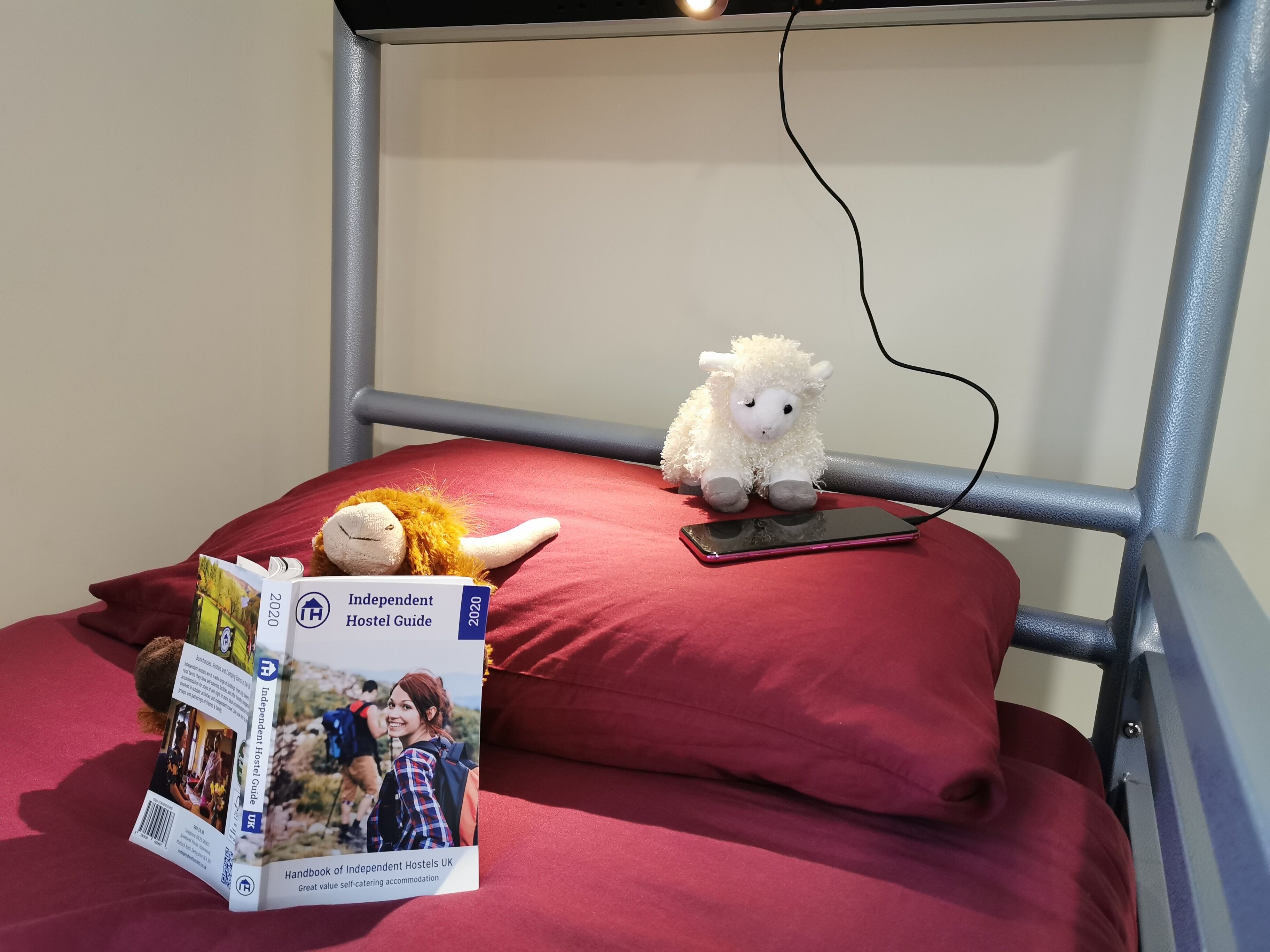 Uma cama com um travesseiro vermelho, um carneirinho de pelúcia, um smartphone e um guia de hostel.
